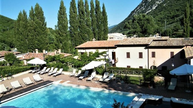Piscina com espreguiçadeiras e guarda-sóis, cercada por edifícios e árvores.