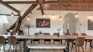 Uma sala de jantar com uma longa mesa de madeira, várias cadeiras, luminárias pendentes e uma pintura de lareira na parede.