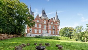 Um edifício grande e histórico, com fachada simétrica, várias torres e um gramado bem cuidado com tocos de madeira espalhados.