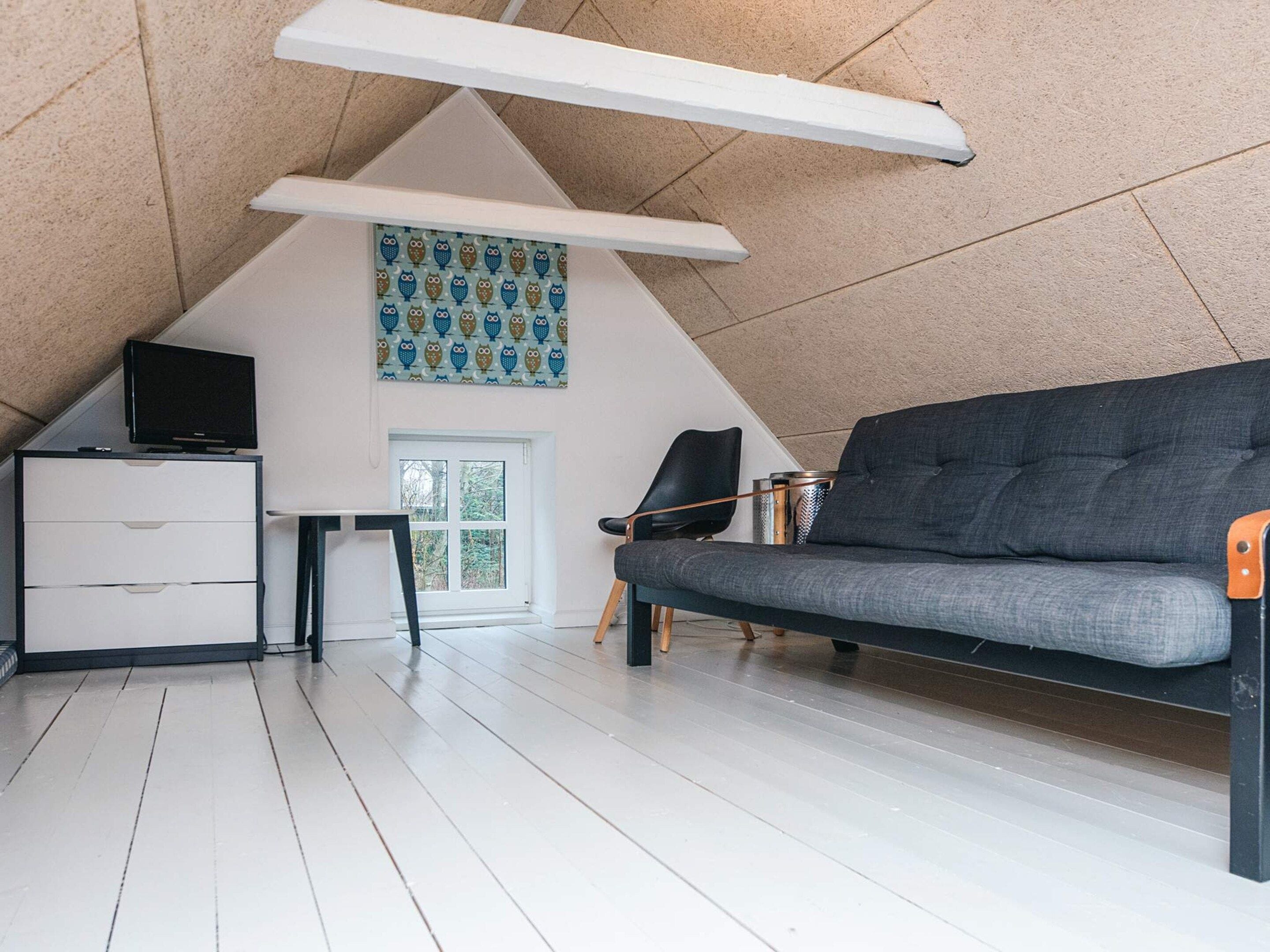 Quarto moderno no estilo loft, com teto inclinado, sofá cinzento, mesa pequena, televisão e janela com vista.