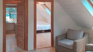 Quarto com teto inclinado, uma cama, uma cadeira de vime e uma escrivaninha com um computador.