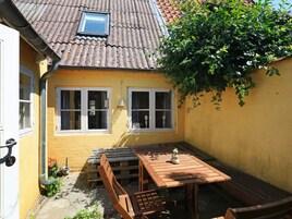 Uma casa amarela com telhado de telhas, duas janelas, uma mesa de madeira com cadeiras e um pequeno jardim.