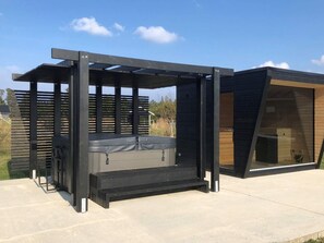 Uma área moderna com hidromassagem ao ar livre, com estrutura de pérgola, deck de madeira e um edifício com telhado inclinado.