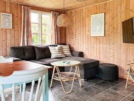 Quarto aconchegante com sofá, mesa redonda e uma cadeira. Há uma janela com cortinas, luminária pendente e um quadro na parede.