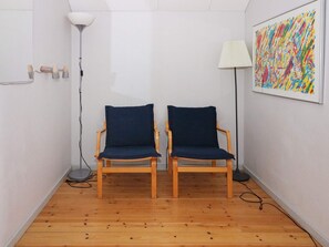 Quarto com duas cadeiras de madeira com almofadas azul-escuro, um abajur de chão branco e um quadro abstrato colorido na parede.