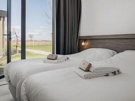 Quarto de hotel moderno com duas camas, uma janela ampla com vista para um campo e cabeceira de madeira com luminária.