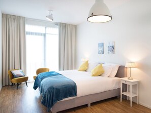 Quarto de hotel com uma cama grande, duas luminárias de cabeceira, uma cadeira e uma janela com cortinas.