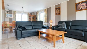 Uma sala de estar com dois sofás pretos, uma mesa de centro de madeira, uma mesa de jantar com cadeiras e um armário.
