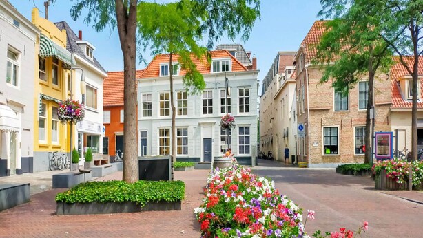 Rua com arquitetura europeia tradicional, canteiros de flores e céu claro.