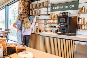 Interior de uma cafeteria com uma máquina de café, prateleiras com sacas de café e um aviso que diz 'Café para levar'.