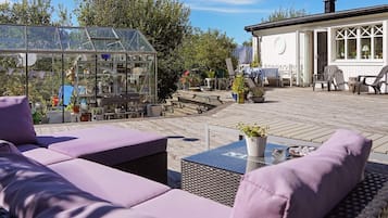 Área de estar ao ar livre coberta, com estrutura tipo estufa de vidro, deck de madeira e mobiliário de lounge.