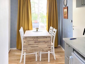 Uma sala de jantar com mesa de madeira e cadeiras brancas, uma janela com cortinas, um relógio e uma área de cozinha com armários e bancada.