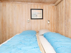 Quarto com paredes de madeira, duas camas com roupa de cama azul, um quadro na parede e duas luminárias fixadas na parede.