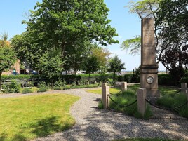 Um monumento comemorativo com inscrições, rodeado por um caminho de cascalho e vegetação.