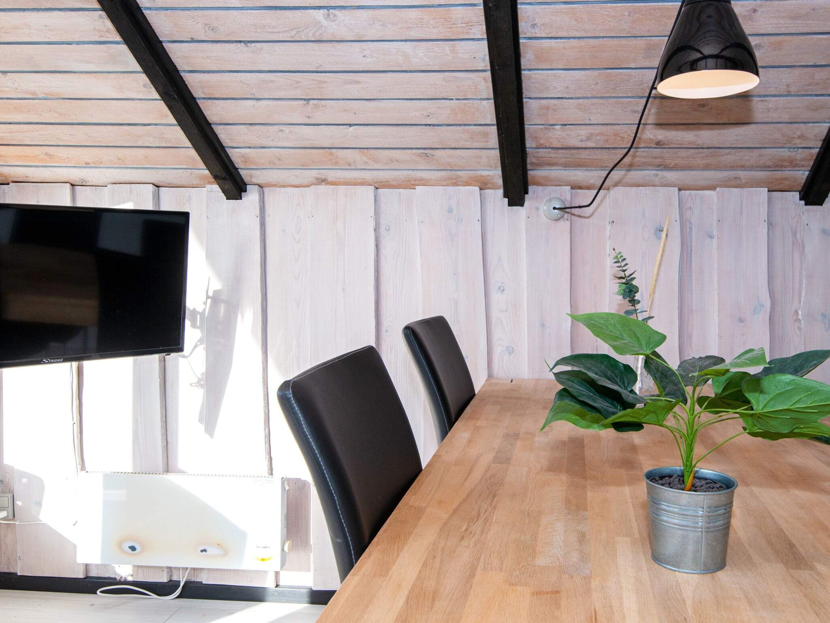 Uma sala de conferências com uma mesa de madeira, cadeiras pretas, uma planta em vaso, uma televisão e uma luminária pendente.