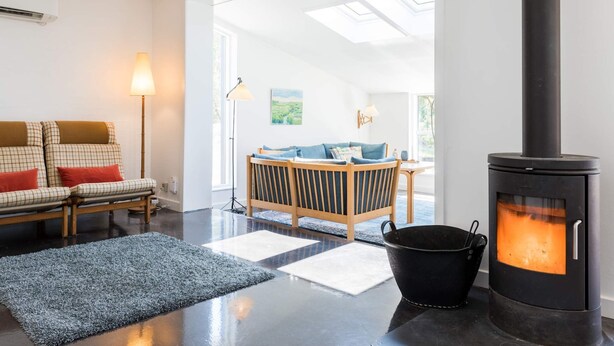 Uma sala de estar moderna com uma lareira a lenha preta, um sofá com almofadas xadrez e vista para um jardim através de claraboias.
