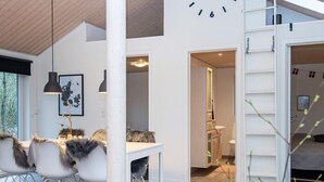 Um interior moderno com área de jantar, com cadeiras brancas e mantas de pele, uma mesa branca e iluminação pendente. Há um relógio visível na parede e uma escada que leva a um andar superior.