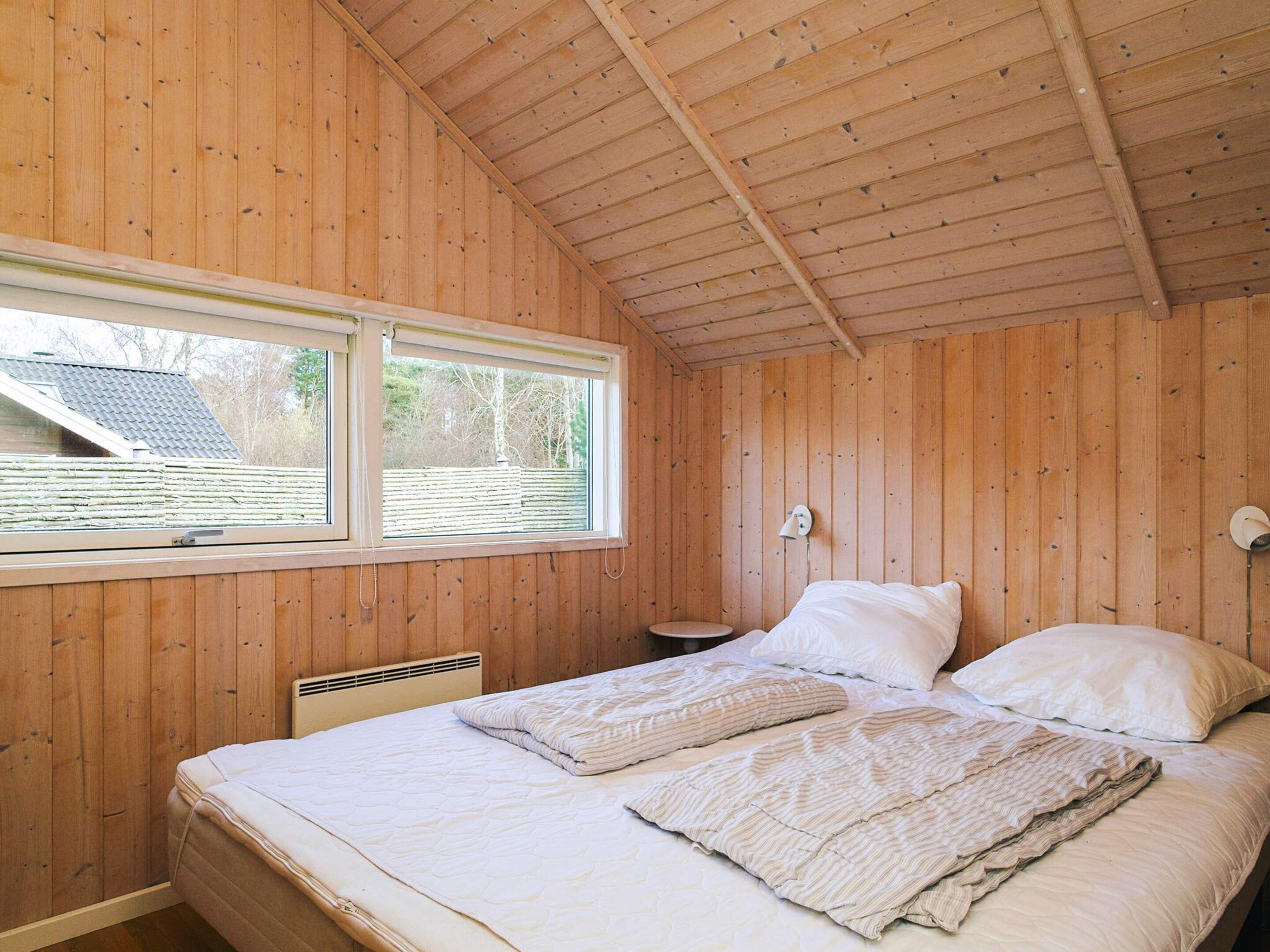 Quarto com paredes de madeira, cama com roupa de cama branca, uma janela e luminária fixada na parede.