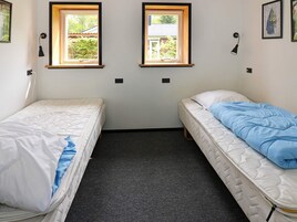 Quarto pequeno com duas camas de solteiro, piso coberto por carpete e duas janelas com moldura de madeira.