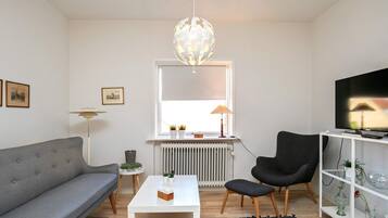 Uma sala de estar moderna com um sofá cinzento, uma mesa de centro branca, uma poltrona preta, uma TV sobre um suporte branco e uma luminária esférica diferenciada.