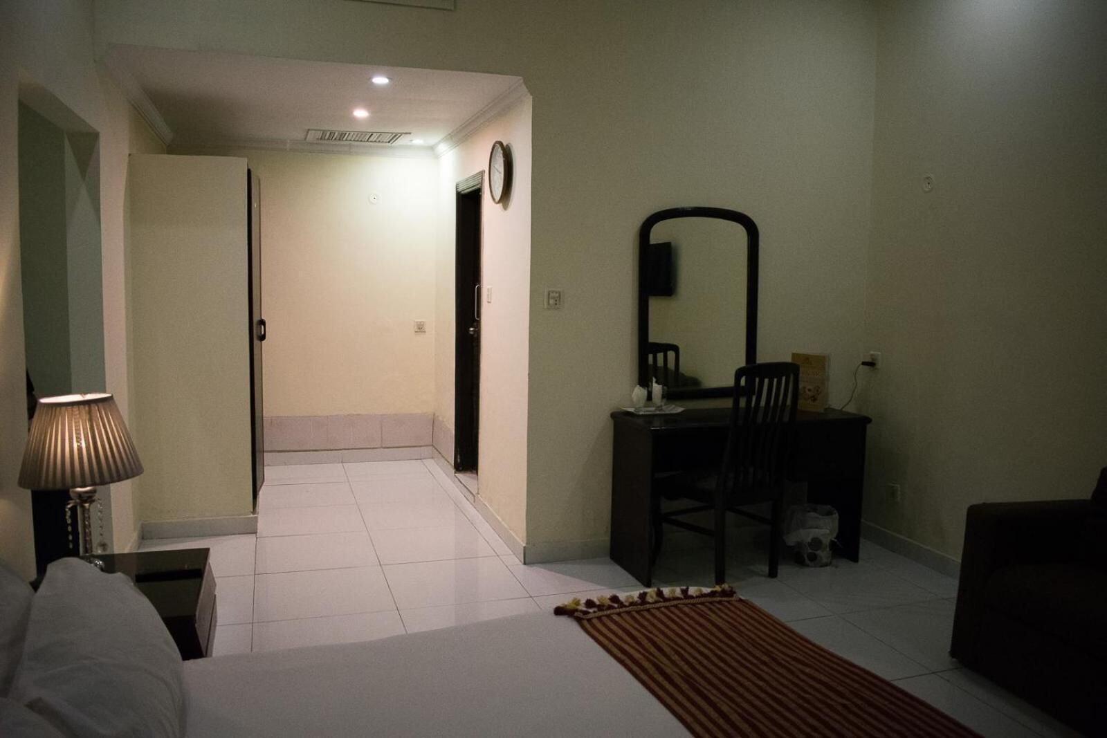 Quarto de hotel com cama, escrivaninha com cadeira, luminária e relógio na parede.