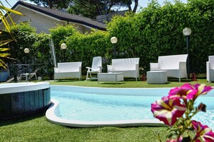 Uma área de piscina com móveis de vime branco, uma banheira de hidromassagem e um gramado bem cuidado.