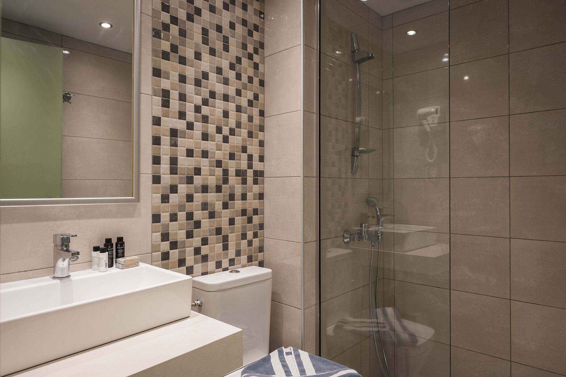 Um banheiro moderno com um revestimento de azulejos xadrez, box de vidro para o chuveiro e pia branca com produtos de higiene.