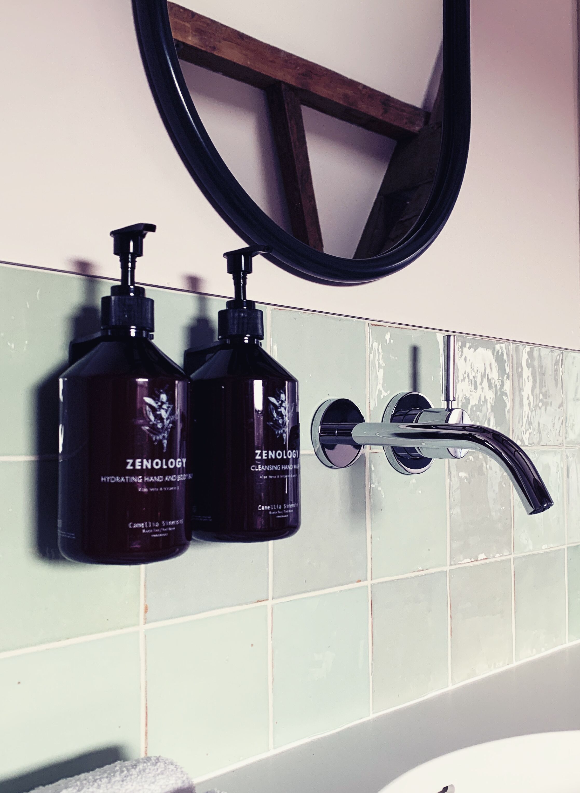 Casa de banho com lavatório branco, duas embalagens pretas de gel de limpeza Zenology, torneira cromada e espelho com moldura de madeira.