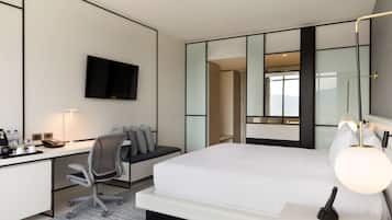 Quarto de hotel moderno com uma cama grande, uma escrivaninha com computador, uma cadeira, TV fixada na parede e uma janela ampla com vista.