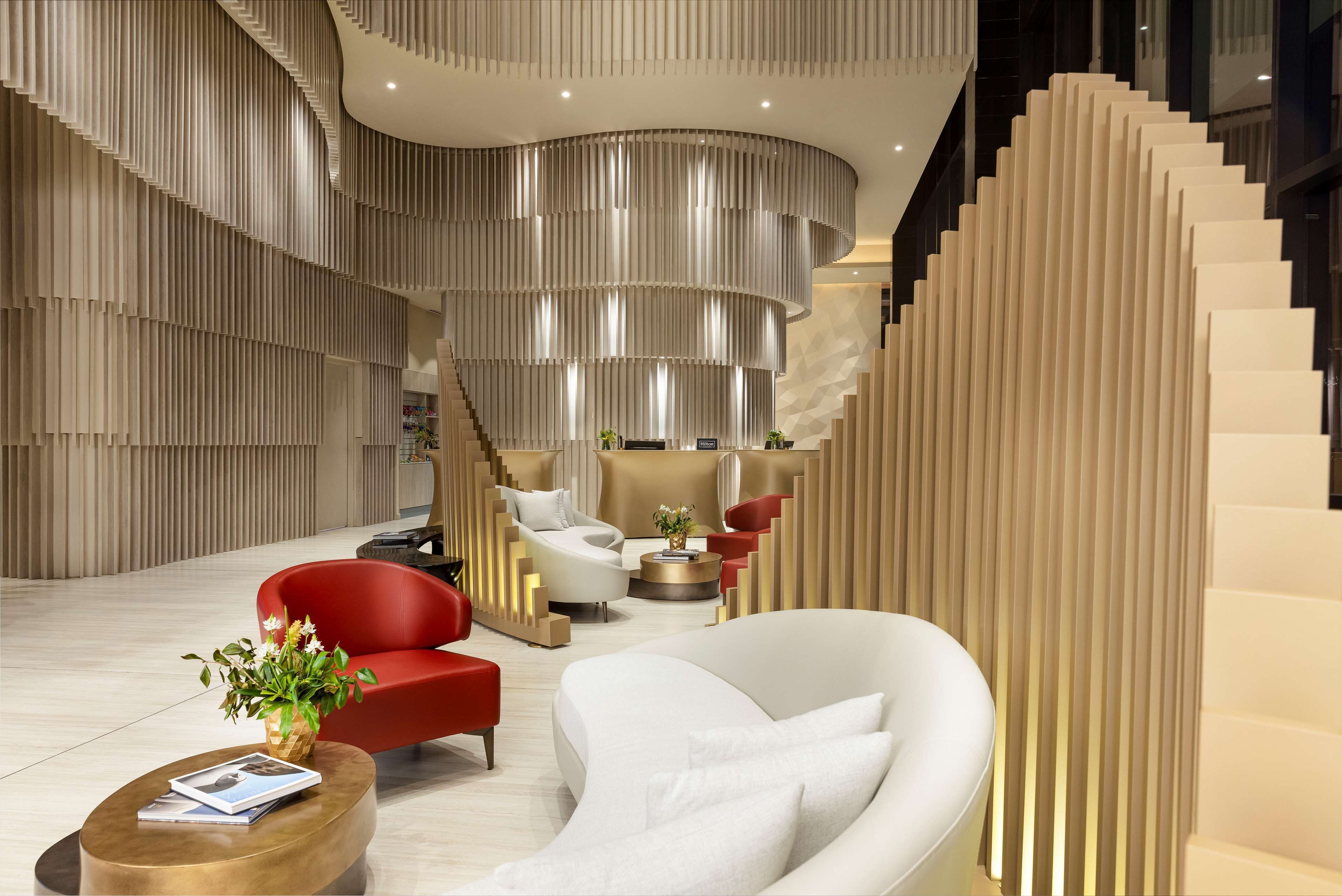 Um lobby moderno com uma escada curva, uma poltrona vermelha, um sofá branco e uma mesa de centro de madeira.