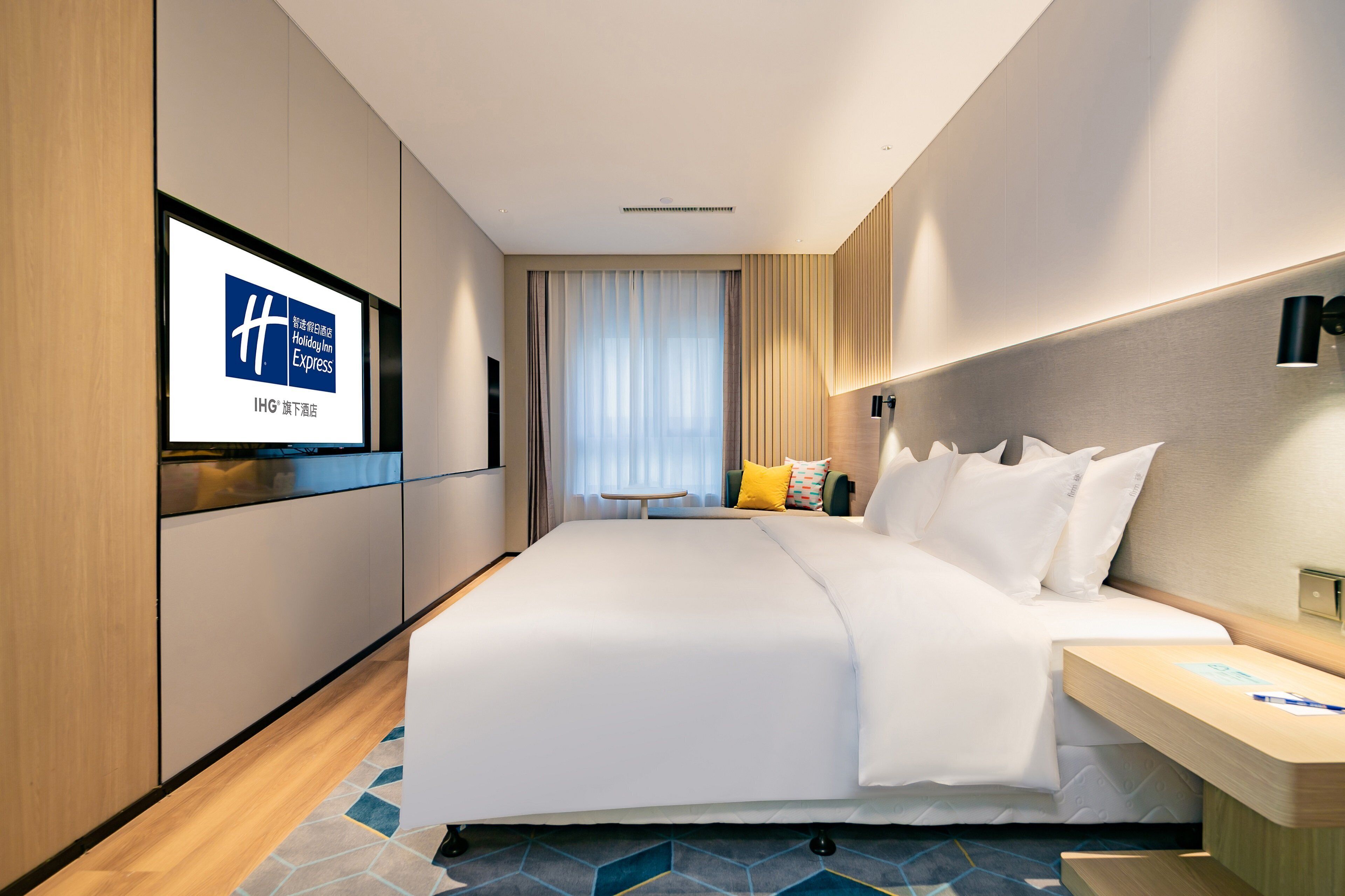 Quarto de hotel com cama, mesa de cabeceira, televisão exibindo o nome do hotel, janela com cortinas e uma pequena mesa com uma cadeira.
