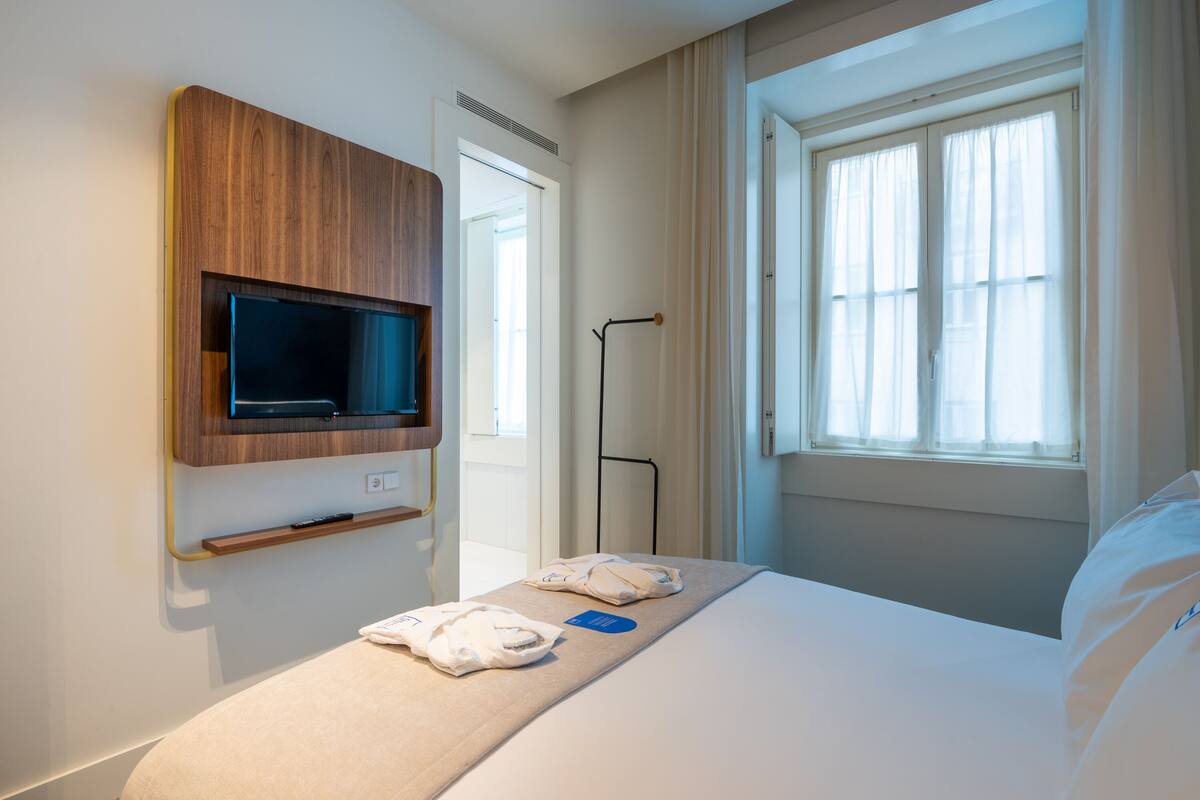 Quarto de hotel com uma cama, televisão fixada em um painel de madeira, mesa de cabeceira e janela com cortinas.