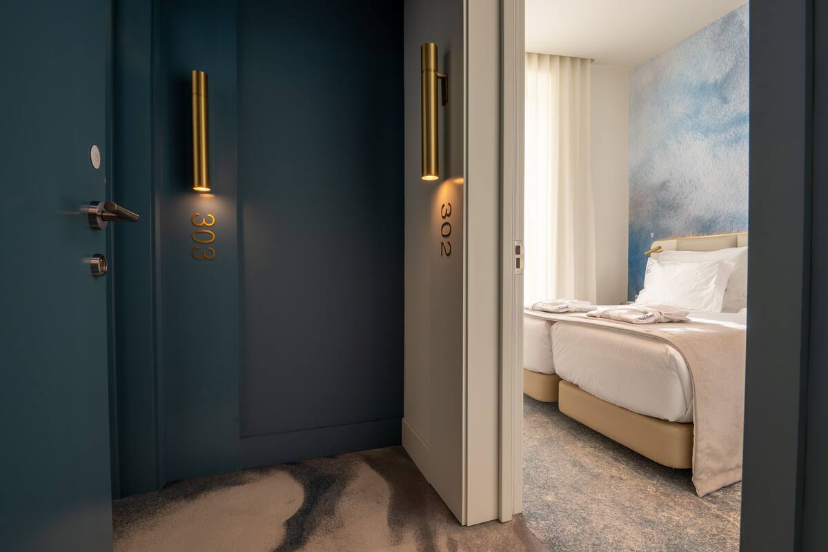 Quarto de hotel moderno com parede azul de destaque, cama com roupa de cama branca e porta com o número 302.