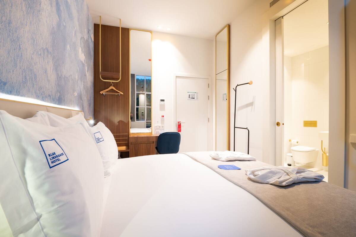 Quarto de hotel com cama, escrivaninha, cadeira, espelho e banheiro visível por uma porta aberta.