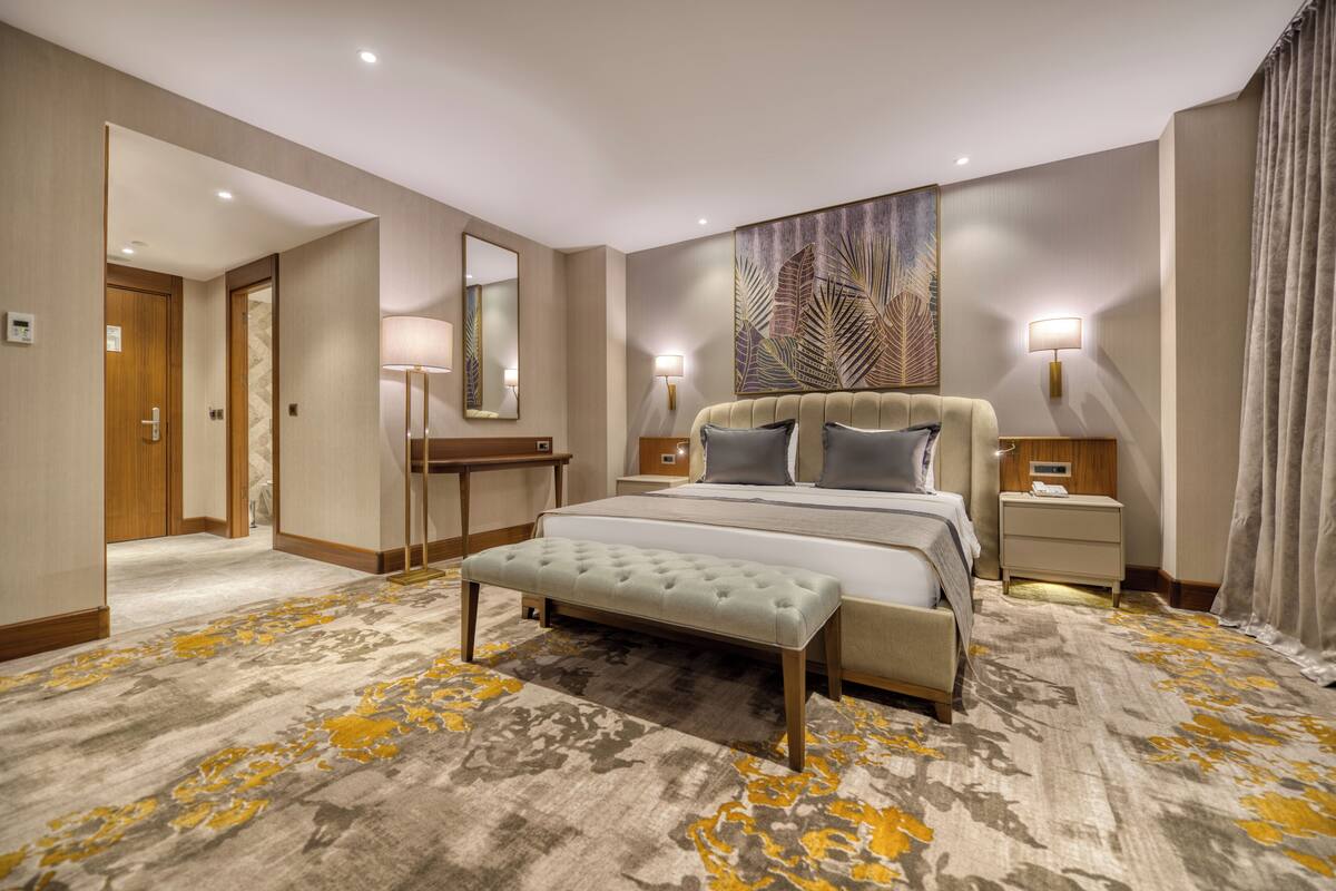 Quarto de hotel com uma cama grande, um banquinho, uma mesa de cabeceira, um espelho e uma luminária fixada na parede.