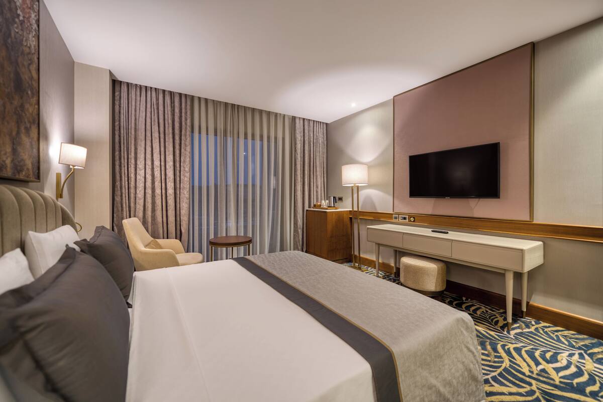 Quarto de hotel com uma cama grande, escrivaninha, cadeira, televisão e janela com cortinas.