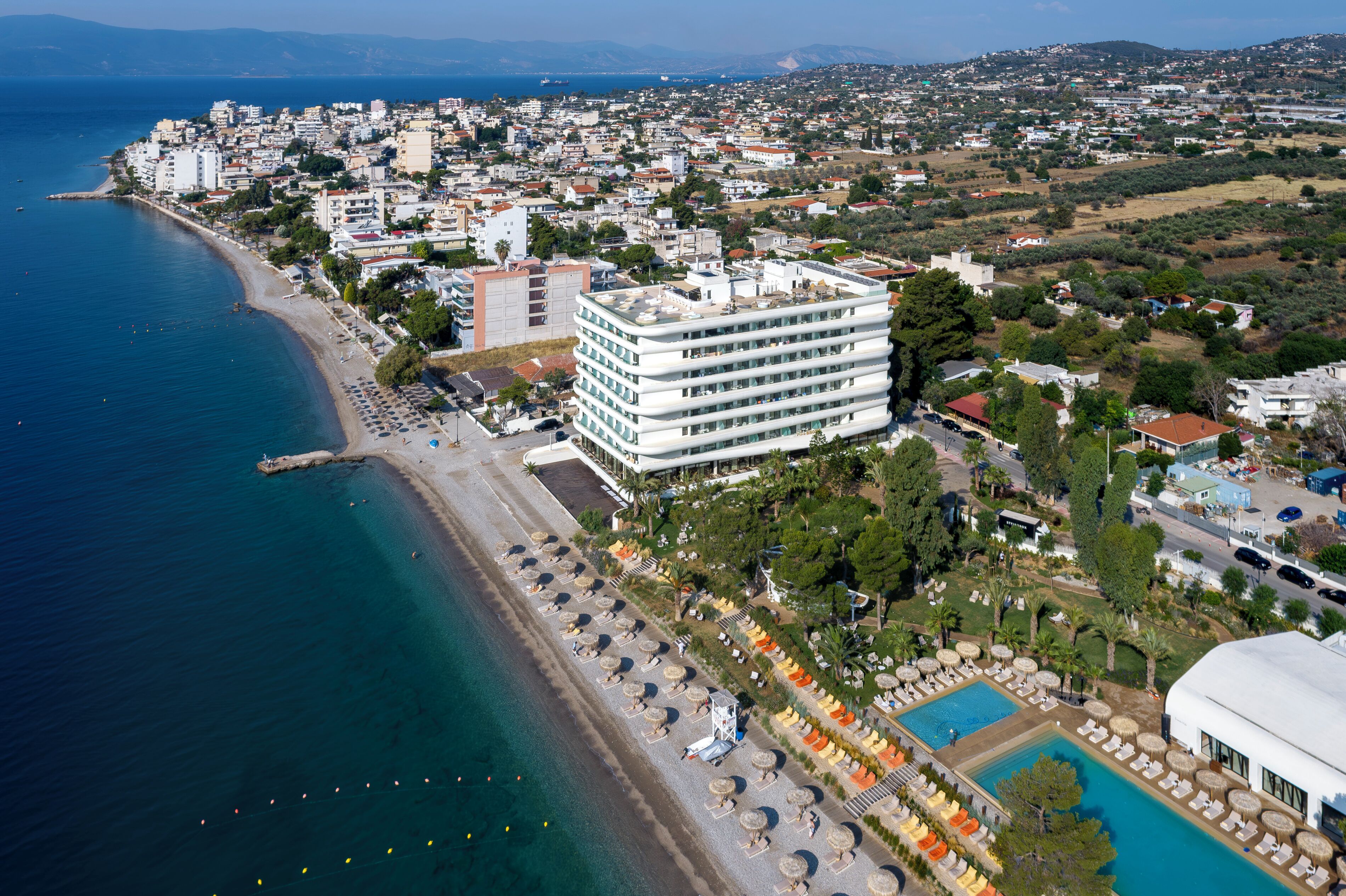 Vista aérea de um hotel à beira-mar, com praia, piscina e um edifício alto com vários andares.