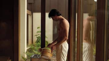 Um homem numa cabana de madeira, segurando um balde, com uma sauna visível através das portas de vidro.