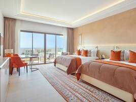 Quarto de hotel com duas camas, uma varanda, uma escrivaninha e vista para o mar.