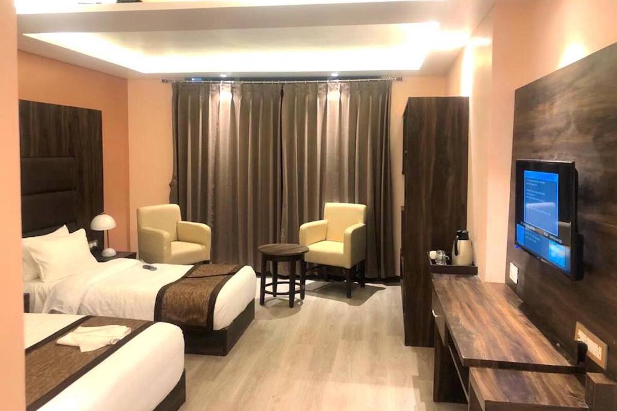Quarto de hotel com uma cama, duas poltronas, uma mesa pequena, televisão e uma janela grande com cortinas.