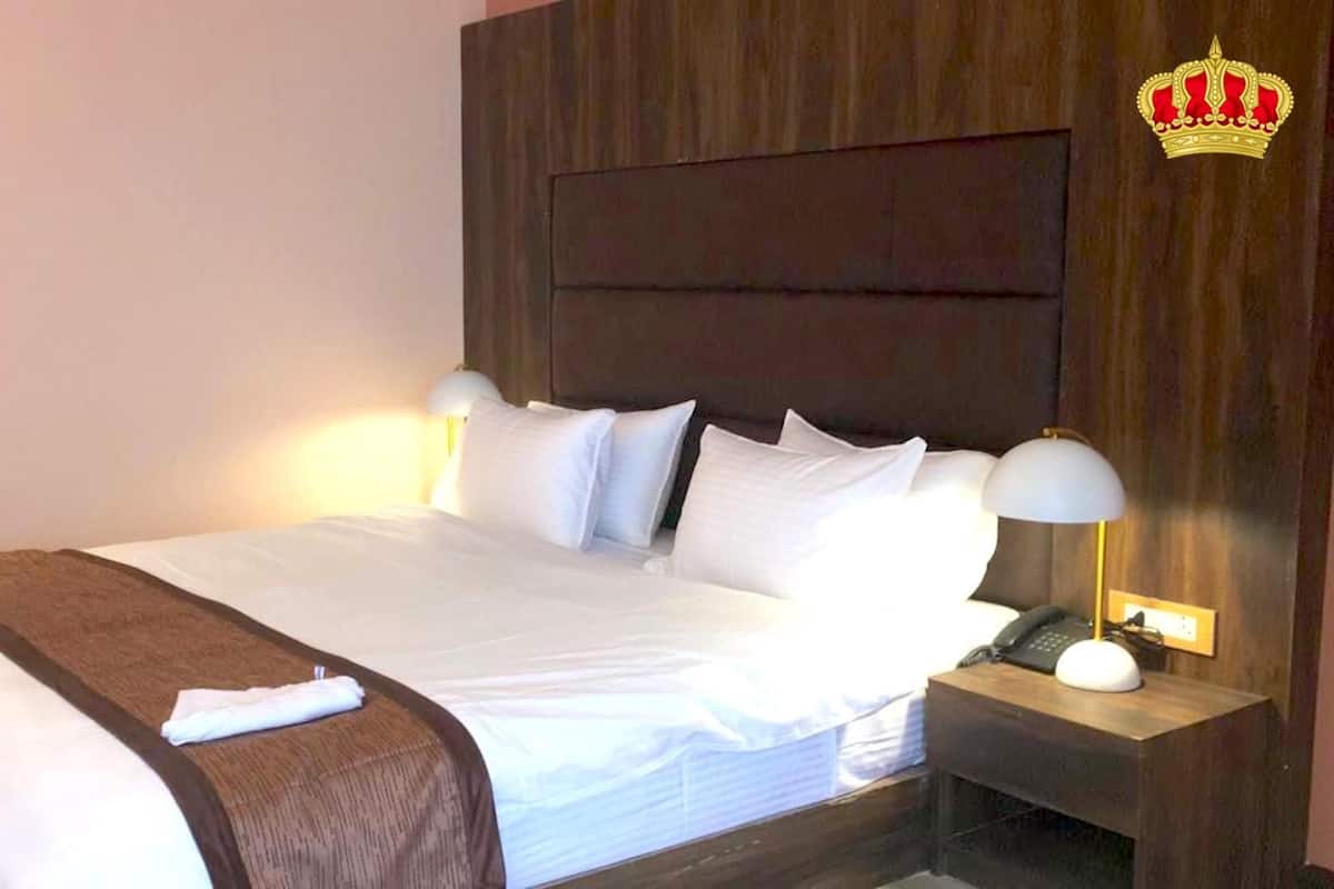 Quarto de hotel com uma cama grande, roupa de cama branca, manta marrom, duas luminárias de cabeceira e uma cabeceira de madeira.