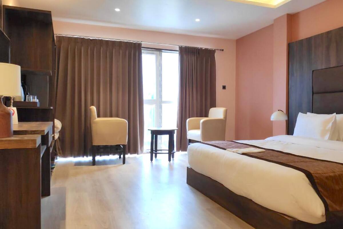 Quarto de hotel com uma cama grande, duas cadeiras, uma escrivaninha com abajur e uma janela com cortinas.