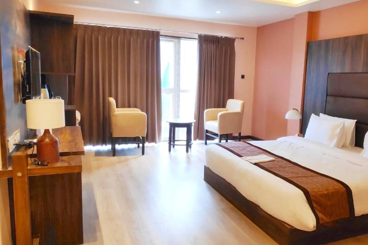 Quarto de hotel com uma cama grande, duas cadeiras, uma escrivaninha com abajur e uma televisão.