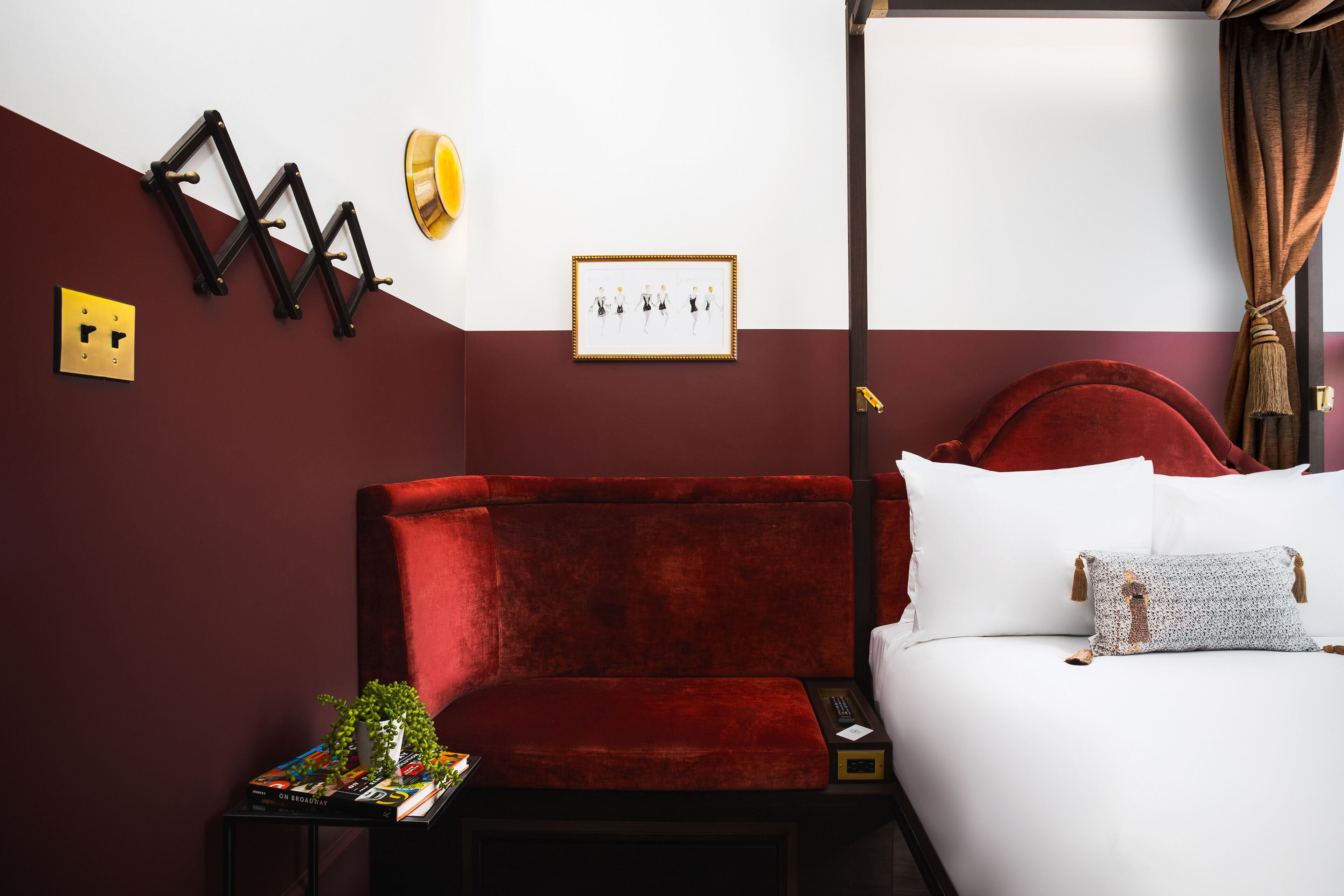 Quarto de hotel com cabeceira de veludo vermelha, cama branca, uma mesinha com planta e livros, e um quadro na parede.