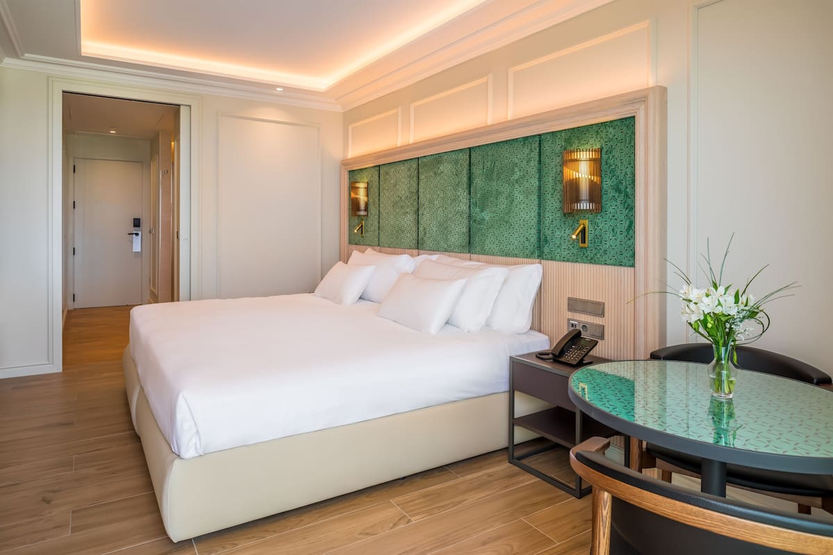 Quarto de hotel com uma cama grande, uma mesa redonda de vidro e uma cabeceira verde com padrão.