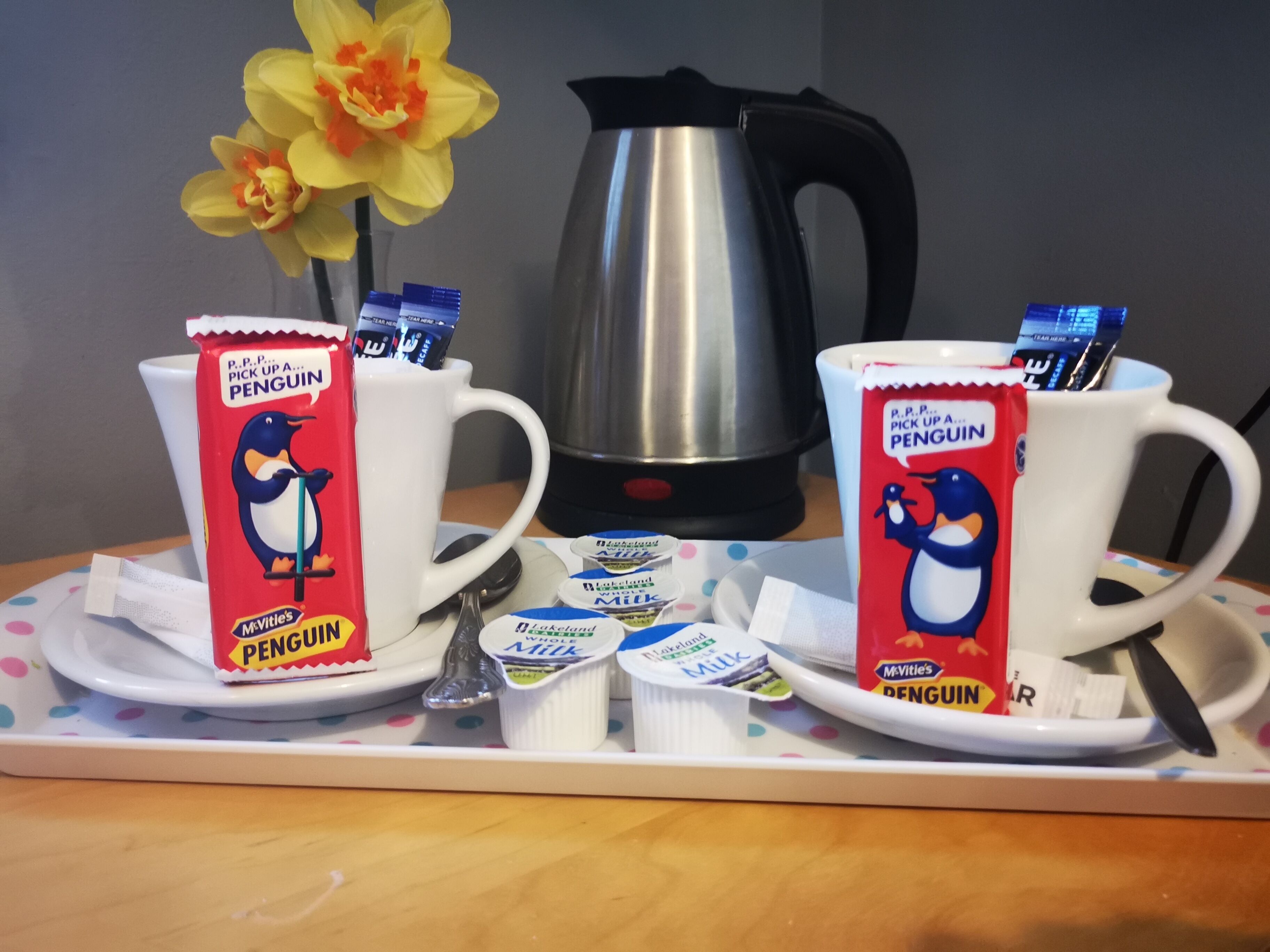 Três canecas com desenhos de pinguins, uma chaleira e alguns produtos lácteos sobre uma bandeja.