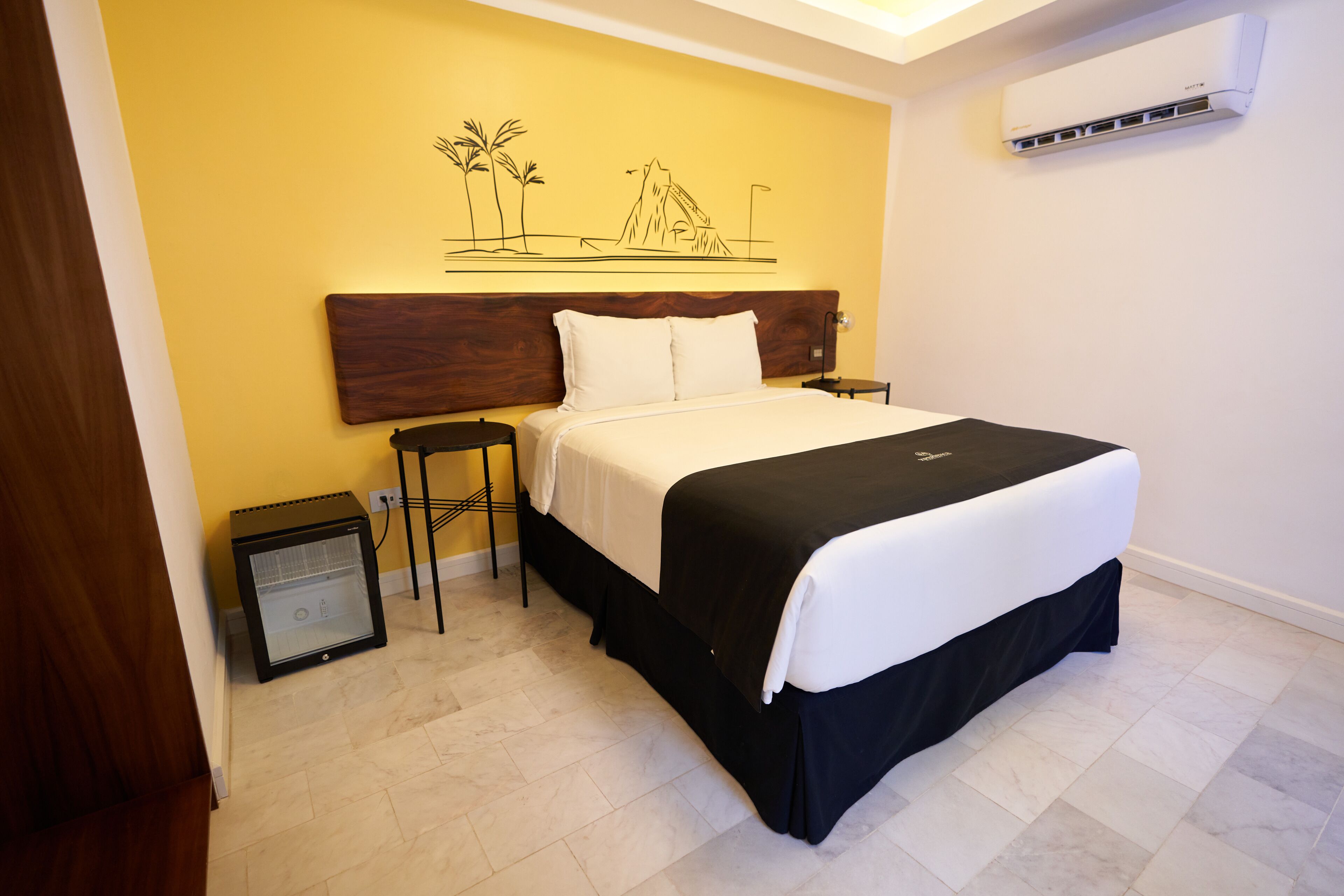 Quarto de hotel com cama, mesa de cabeceira, mesinha e ar condicionado, além de um mural na parede.