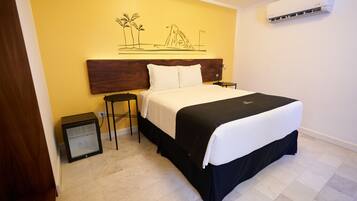 Quarto de hotel com cama, mesa de cabeceira, mesinha e ar condicionado, além de um mural na parede.