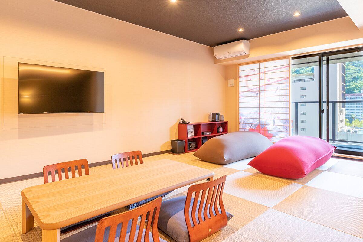Uma sala de estar moderna com uma mesa de jantar de madeira, cadeiras, uma TV de tela plana, uma estante vermelha e uma grande janela com vista para edifícios.