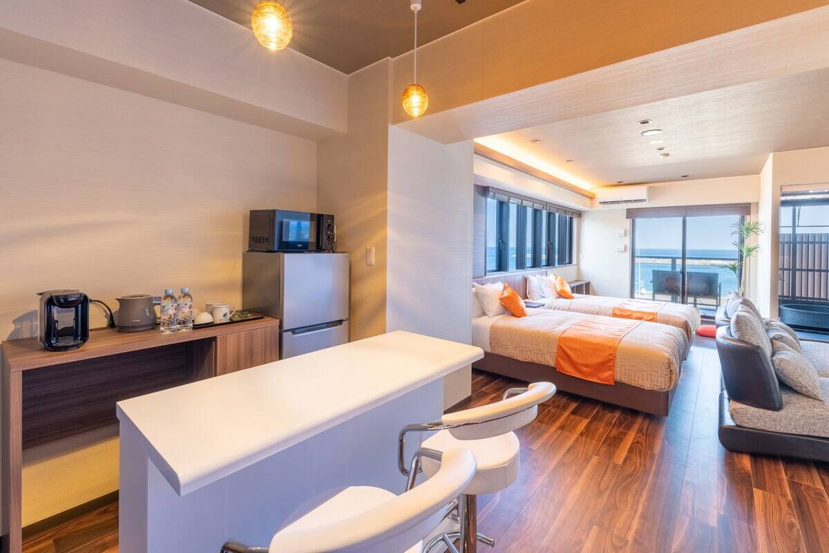 Quarto de hotel moderno com cozinha compacta, uma cama grande com roupa de cama laranja, área de estar com sofá e poltronas, e varanda com vista.